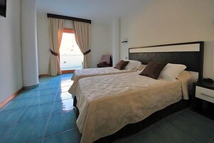 Quarto de hotel com duas camas, mesa de cabeceira, janela com cortinas e chão de azulejos.
