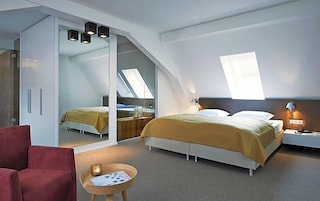 Quarto moderno com cama, mesas de cabeceira, uma cadeira vermelha e uma pequena mesa com uma vela.