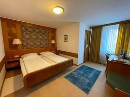 Quarto de hotel com cama de casal, luminárias de cabeceira, escrivaninha com cadeira, mesinha, janela com cortinas e uma obra de arte na parede.