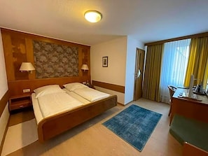 Quarto de hotel com cama de casal, luminárias de cabeceira, escrivaninha com cadeira, mesinha, janela com cortinas e uma obra de arte na parede.