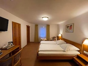 Quarto de hotel com cama, mesa de cabeceira, luminária, televisão e vista para o corredor.