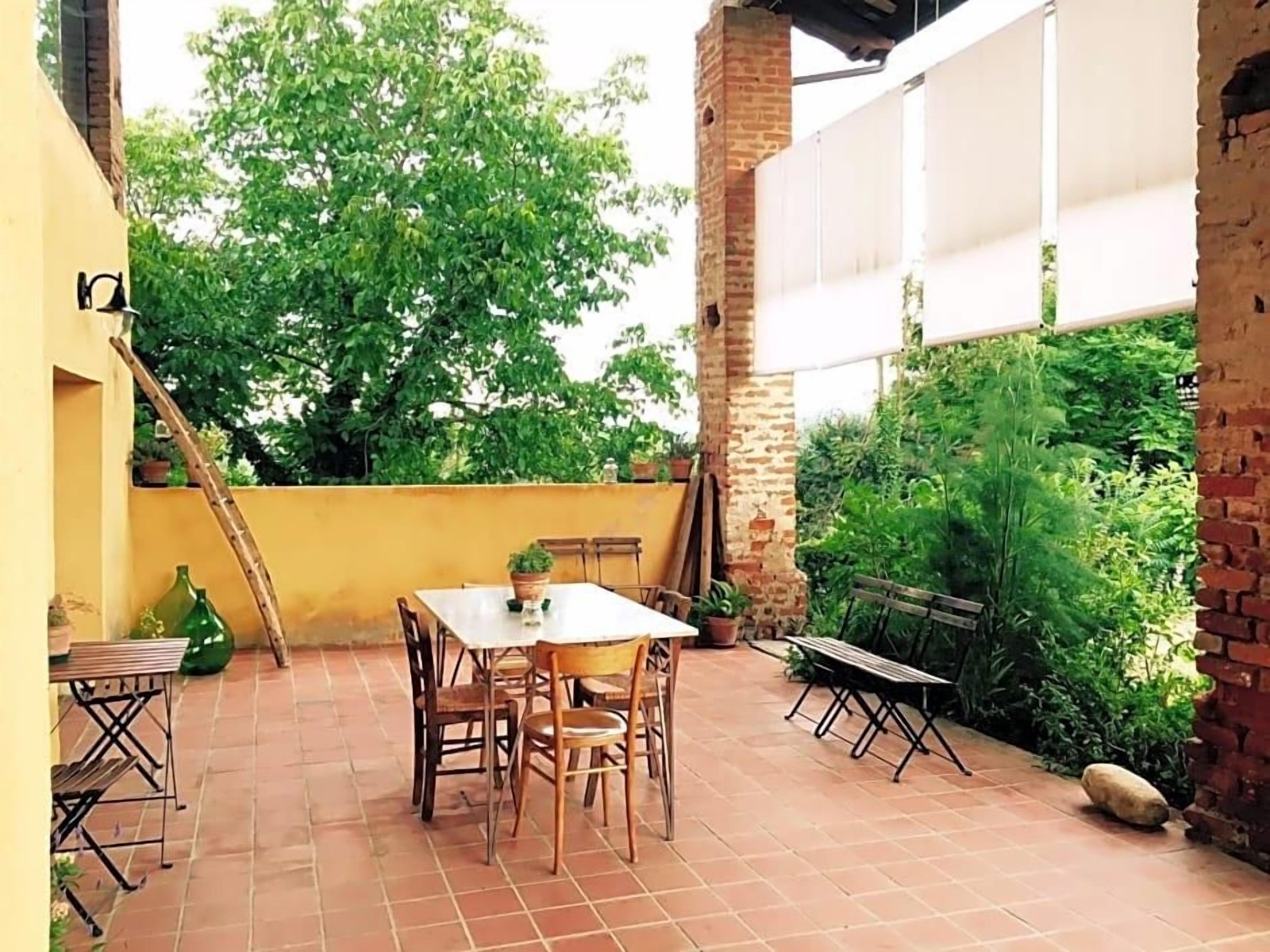 Pátio externo com mesa e cadeiras, cercado por vegetação e uma parede de tijolos.