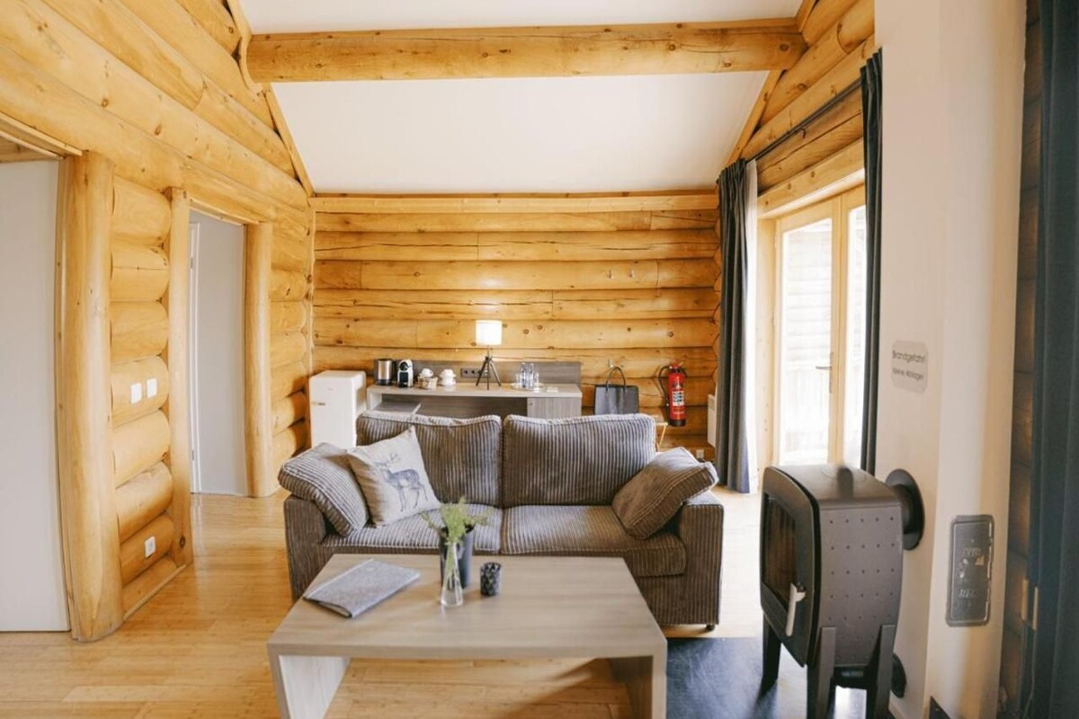 Interior de cabana de troncos com sala de estar, sofá, mesa de centro, lareira e cozinha.