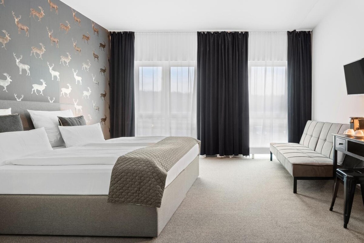Quarto de hotel com cama, escrivaninha, cadeira, televisão e janela com cortinas.