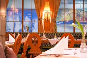 Uma sala de jantar com cadeiras de madeira, uma mesa posta com toalha branca e vista através de grandes janelas.