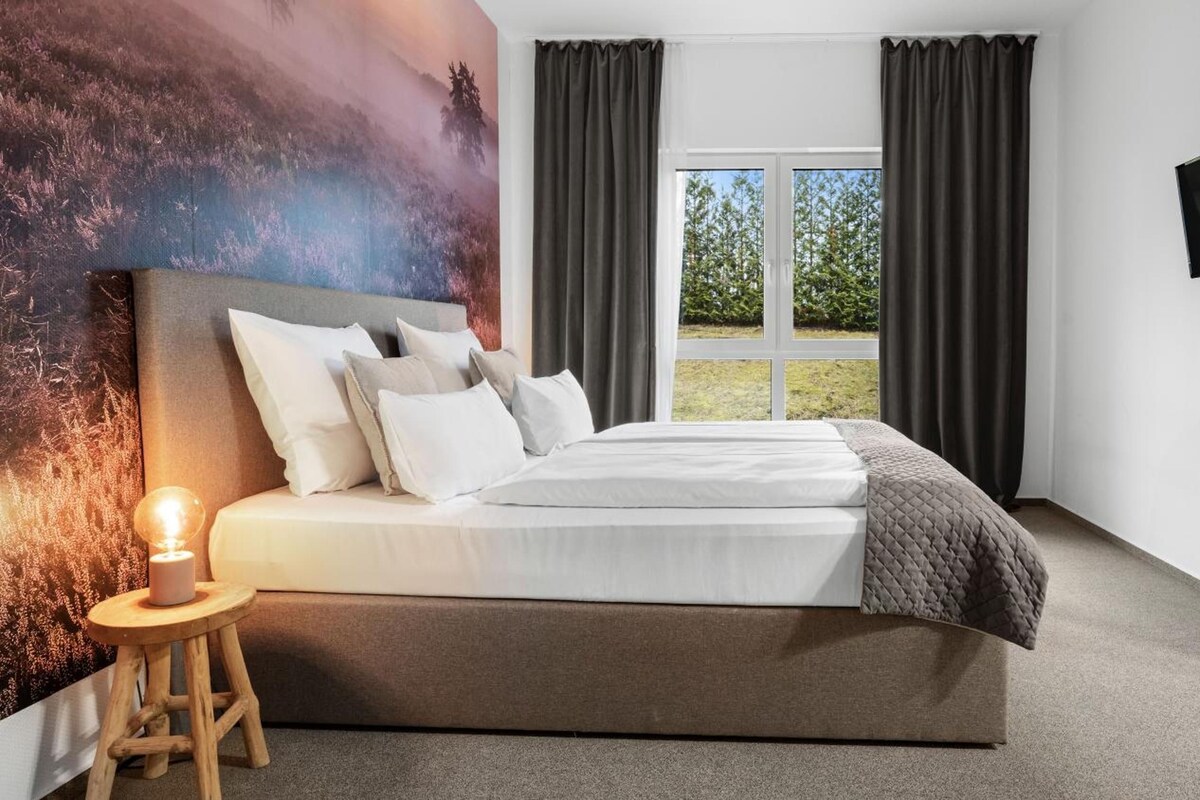 Quarto moderno com uma cama grande, mesa de cabeceira, TV na parede e vista para o exterior.