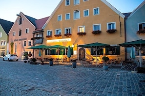 Uma rua tradicional europeia com um café chamado 'Winkler' e um prédio com a placa 'Bauern Brauerei'.