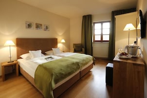 Quarto de hotel com cama grande, duas luminárias de cabeceira, mesa de cabeceira de madeira, uma mesinha com um balde e uma janela com cortinas.
