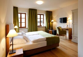 Quarto de hotel com cama, duas luminárias de cabeceira, escrivaninha com cadeira, televisão e guarda-roupa.