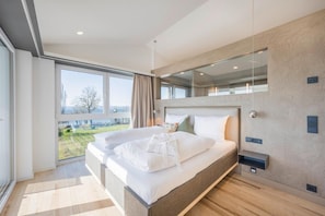 Quarto moderno com cama grande, vista para o exterior e espelho embutido.