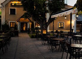 Um pátio de restaurante ao ar livre com mesas e cadeiras, um edifício com o letreiro 'RESTAURANT' e uma árvore no centro.