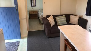 Espaço de estar compacto com sofá, mesa pequena, cama com manta listrada e porta que dá para outro cômodo.