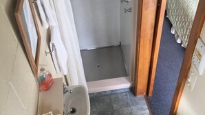 Casa de banho com chuveiro, pia e uma cama visível através de uma porta aberta.