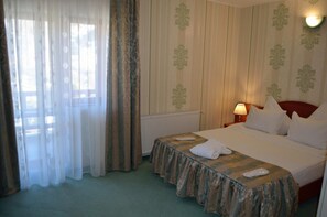 Quarto de hotel com cama, mesa de cabeceira com abajur e janela com cortinas.