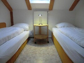 Quarto pequeno e simples com duas camas de solteiro, uma mesa e janela com persianas.