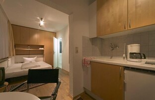 Quarto de hotel compacto com cama, pequena escrivaninha, cadeira, cozinha compacta com cafeteira e armários de madeira.