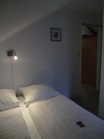 Uma cama de solteiro com roupa de cama branca, luminária fixada na parede e um quadro emoldurado.