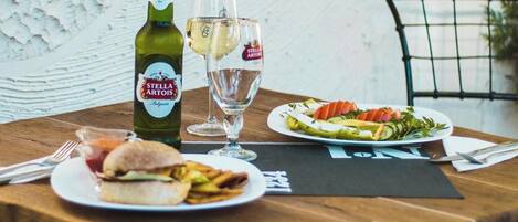 Uma mesa com uma garrafa de Stella Artois, um copo de vinho branco, um sanduíche, uma salada e talheres.