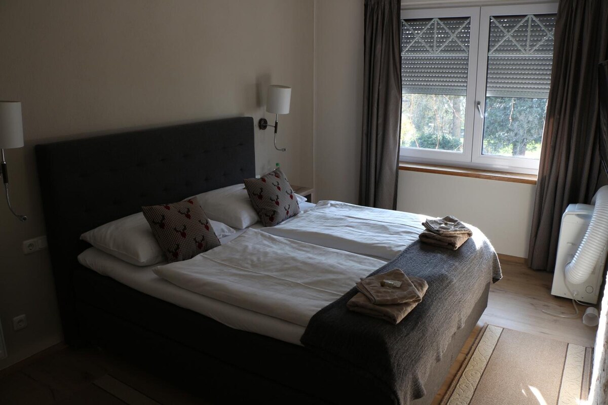 Uma cama de casal com roupa de cama branca, duas almofadas, uma luminária de cabeceira, uma janela com persianas e um aquecedor.