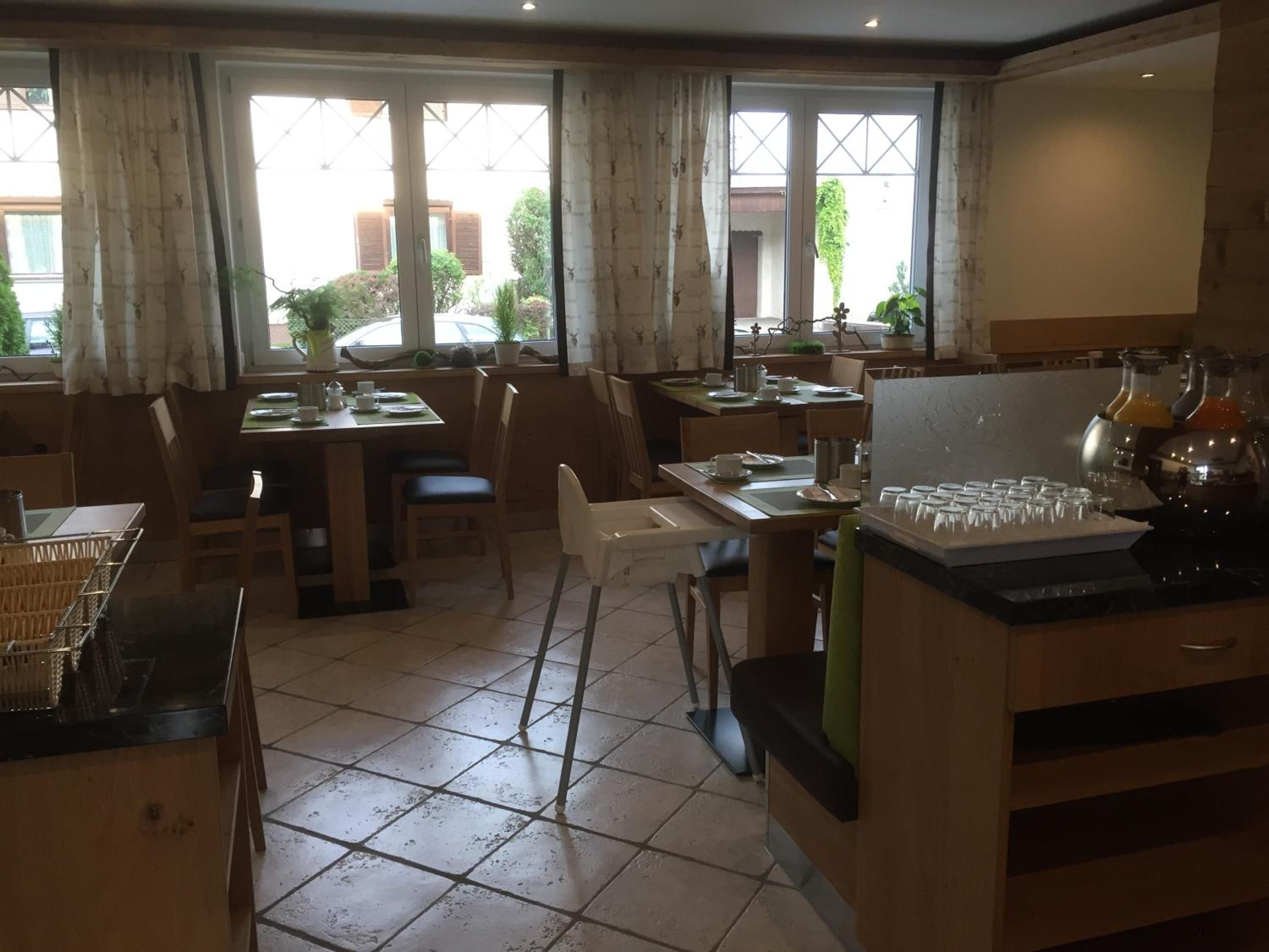 Espaço de refeições com várias mesas e cadeiras, bancada de cozinha com máquina de café e janelas amplas com cortinas.