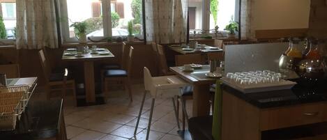 Espaço de refeições com várias mesas e cadeiras, bancada de cozinha com máquina de café e janelas amplas com cortinas.