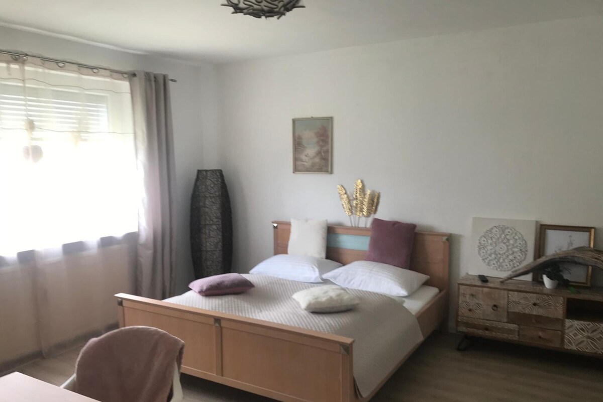 Quarto com cama, poltrona, mesa de cabeceira, cômoda e janela com cortinas.