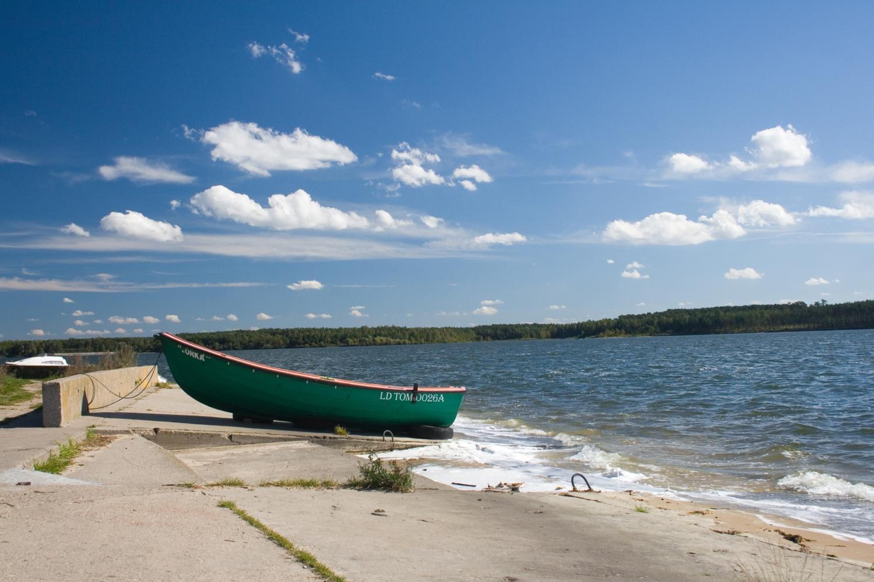 Um barco verde chamado 'LD TOM 0026A' atracado em um cais de concreto, com vista para um lago e uma floresta ao longe.