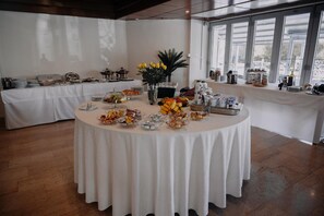 Disposição de buffet com diversos alimentos e bebidas sobre uma mesa redonda, coberta por uma toalha branca.