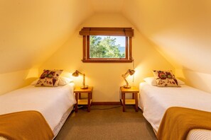 Quarto pequeno e aconchegante com duas camas de solteiro, cada uma com mesa de cabeceira e luminária. Há uma janela sobre as camas, com vista para árvores e uma montanha.