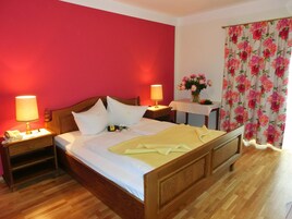 Quarto de hotel com cama de madeira, duas mesas de cabeceira com luminárias, uma mesinha de apoio com telefone, uma pequena mesa com um vaso de flores e cortina florida.