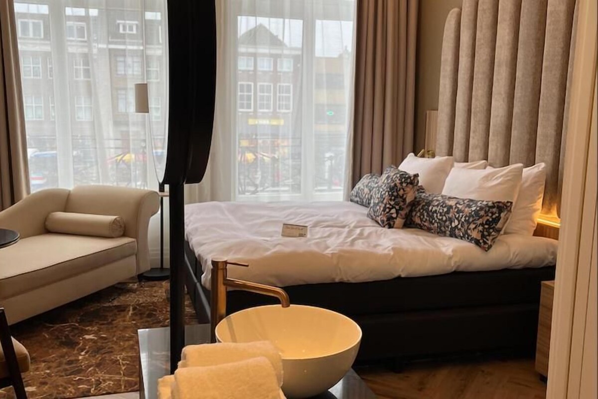 Quarto de hotel com cama, sofá, mesa de cabeceira com uma tigela e vista para edifícios pela janela.