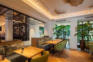 Interior moderno com mesas de madeira e assentos estofados em verde, teto decorativo e janelas amplas com plantas dentro de casa.