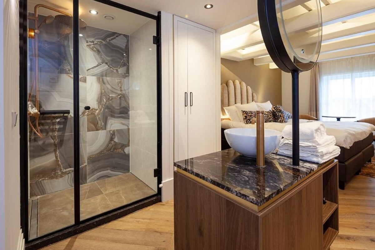Quarto de hotel moderno com chuveiro em box de vidro, toucador de madeira com bancada de mármore e um espelho grande.