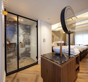 Quarto de hotel moderno com chuveiro em box de vidro, toucador de madeira com bancada de mármore e um espelho grande.