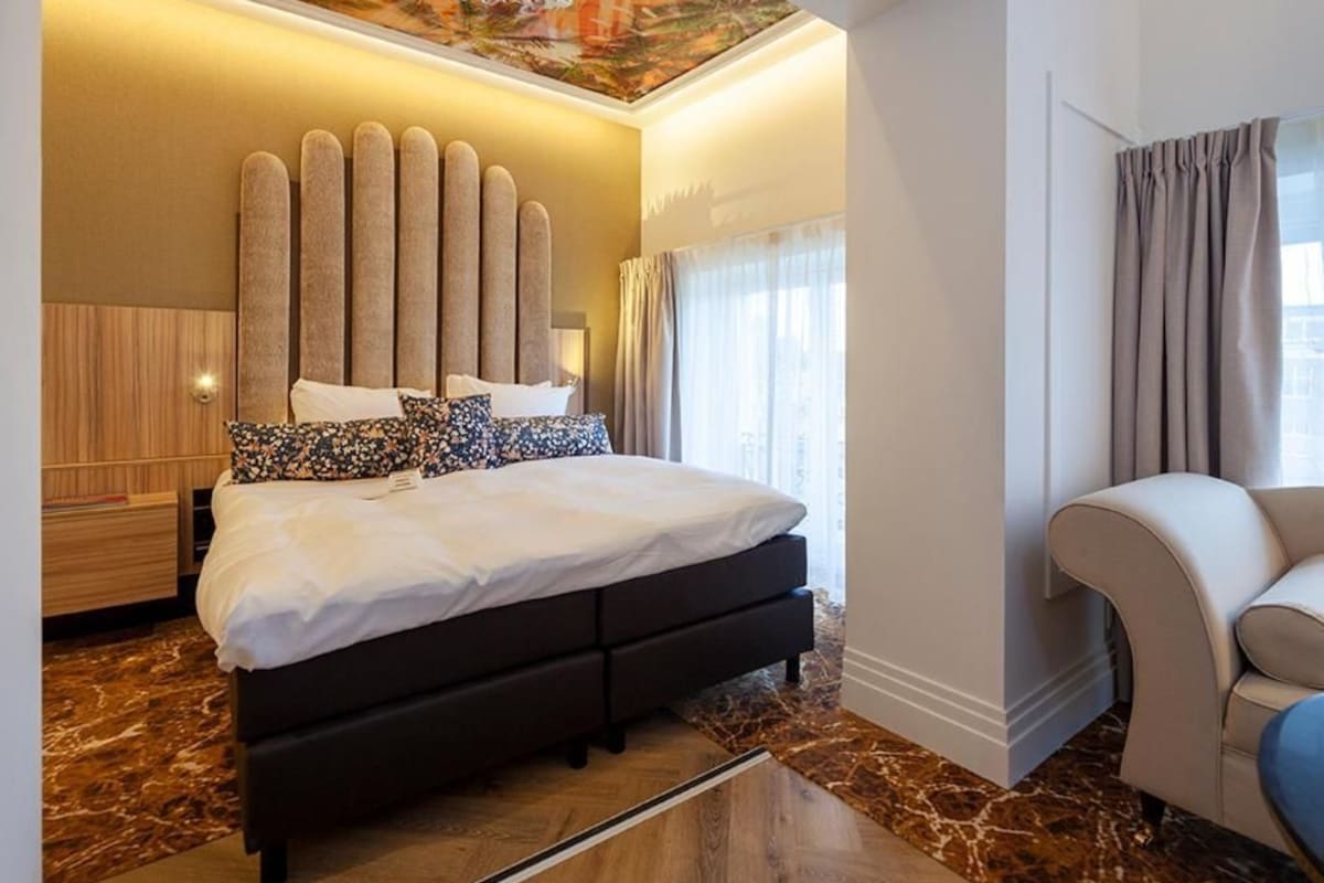 Quarto de hotel moderno com uma cama grande, área de estar e um mural no teto.