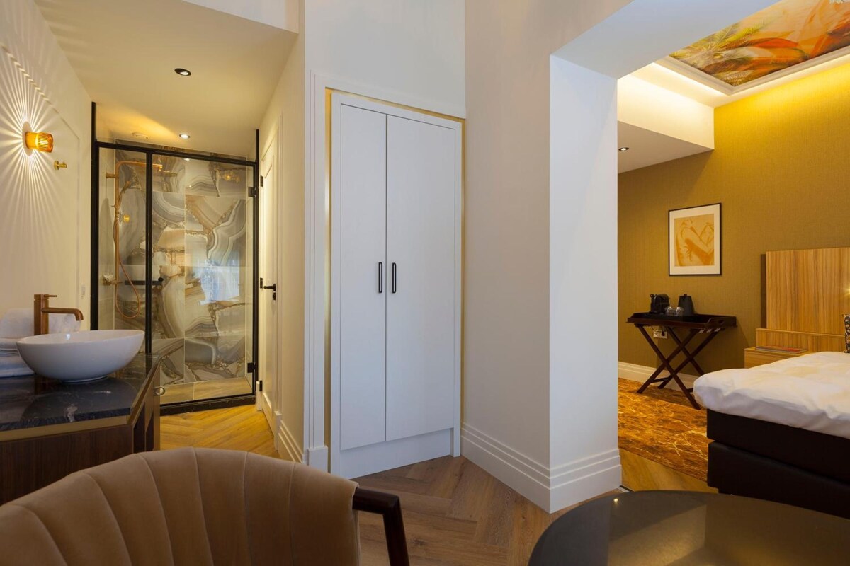 Quarto de hotel com chuveiro em box de vidro, guarda-roupa branco, cama, mesa pequena com luminária e um quadro na parede.