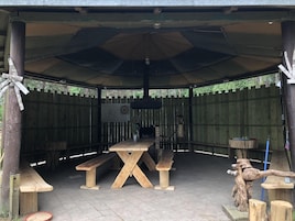 Um pavilhão ao ar livre com mesa e bancos, cercado por uma cerca de madeira.