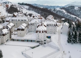 Estância de esqui coberta de neve, com vários edifícios, incluindo um hotel central com uma entrada ampla e um teleférico visível ao fundo.