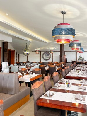 Um restaurante com várias mesas e cadeiras, um candelabro e uma obra de arte pendurada na parede.