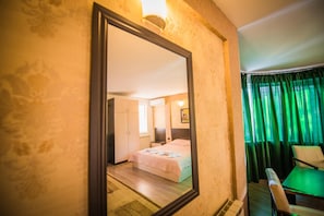Quarto de hotel com cama, escrivaninha e uma cadeira. Há um espelho na parede e uma janela com cortinas.