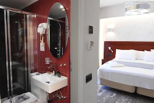 Quarto de hotel com uma cama branca, banheiro com pia e espelho, e chuveiro.