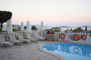 Área de piscina com espreguiçadeiras e guarda-sóis, piscina e vista para o mar e colinas.