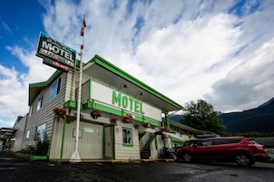 Um motel com fachada verde e branca, uma bandeira no topo e um carro vermelho estacionado em frente.