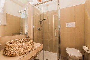 Casa de banho com chuveiro envolto em vidro, lavatório com padrão de pedras arredondadas e sanita com rolo de papel higiénico.