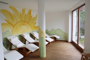 Quarto com piso de madeira, cadeiras de espreguiçadeira brancas, um grande mural de sol na parede e janela com vista para o verde.