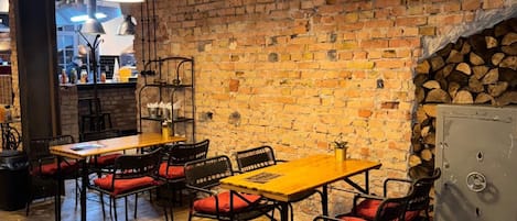 Interior com paredes de tijolo, uma mesa comprida e cadeiras, luminária pendente e lareira com caixa de armazenamento.