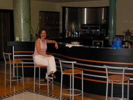 Uma área de bar com uma pessoa sentada, banquetas e um balcão repleto de garrafas.