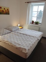 Quarto com duas camas, uma mesa de cabeceira com luminária, uma janela com cortinas e um radiador.