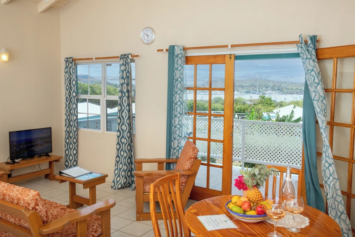 Quarto com televisão, mesa de jantar com frutas e copos de vinho, sofá e vista para a água através de amplas janelas.