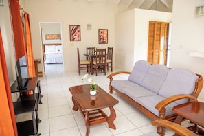 Sala de estar com sofá, mesa de centro de madeira e TV em um suporte. Há uma área de jantar com mesa e cadeiras, e uma cama ao fundo.